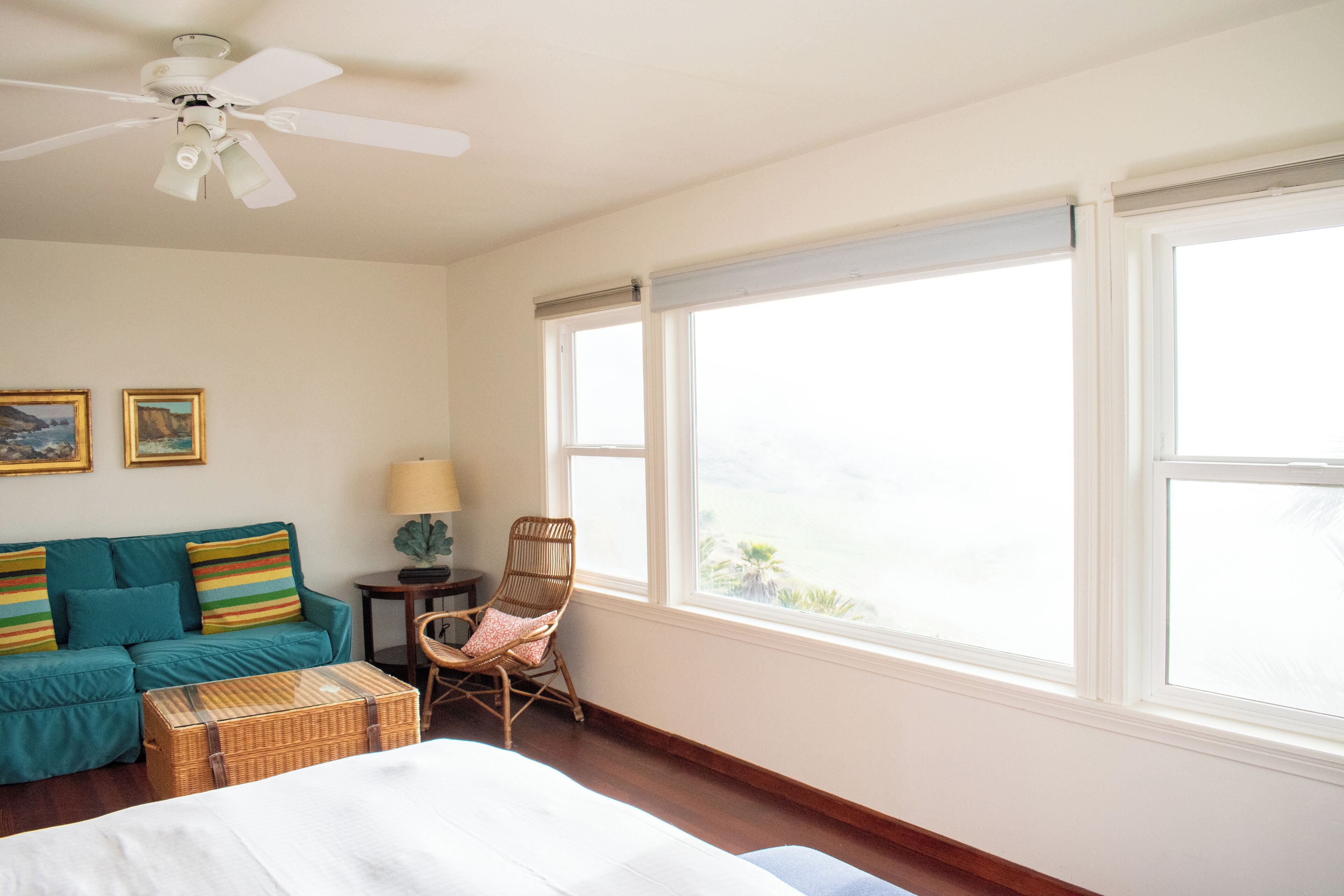 Quarto com uma cama, um sofá verde, uma cadeira de vime, uma mesinha de centro com abajur e grandes janelas com vista.