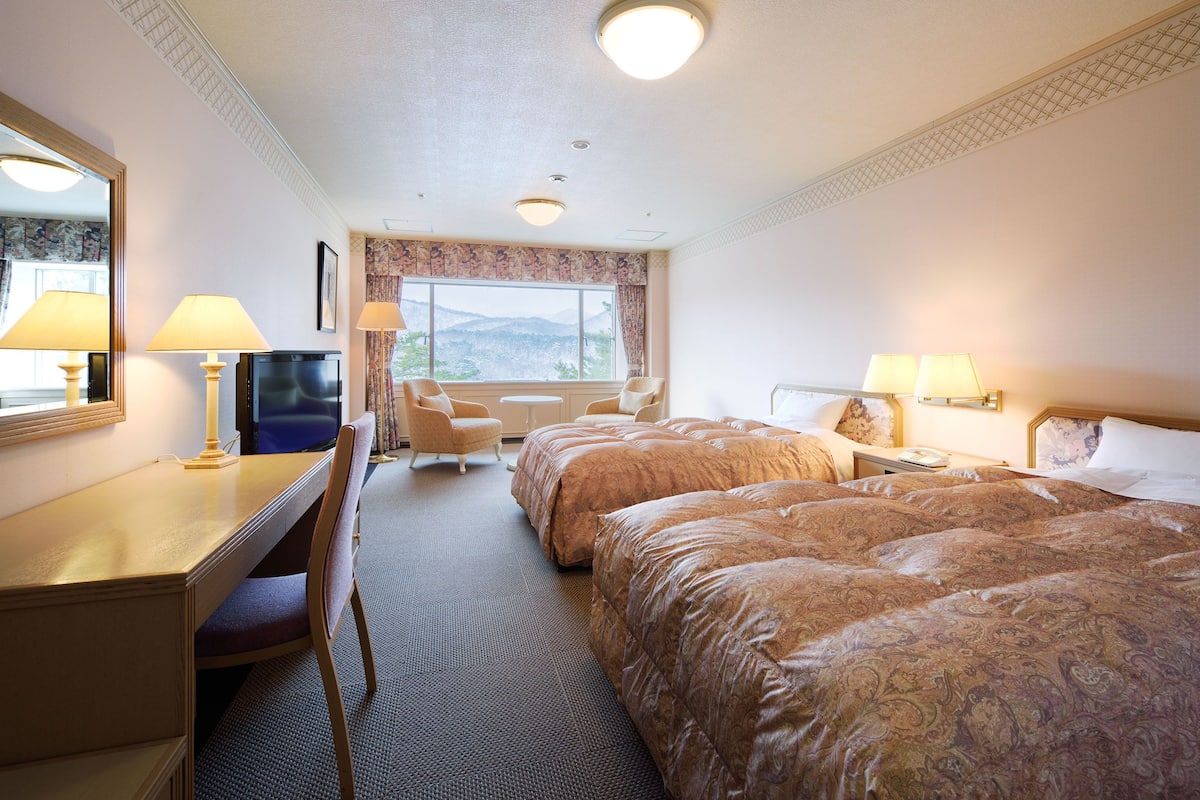 Quarto de hotel com duas camas, uma escrivaninha com cadeira, televisão e janela com vista para as montanhas.