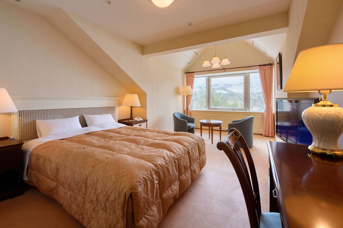 Quarto de hotel com uma cama grande, uma escrivaninha com luminária, duas cadeiras, uma TV e uma janela com vista para o exterior.