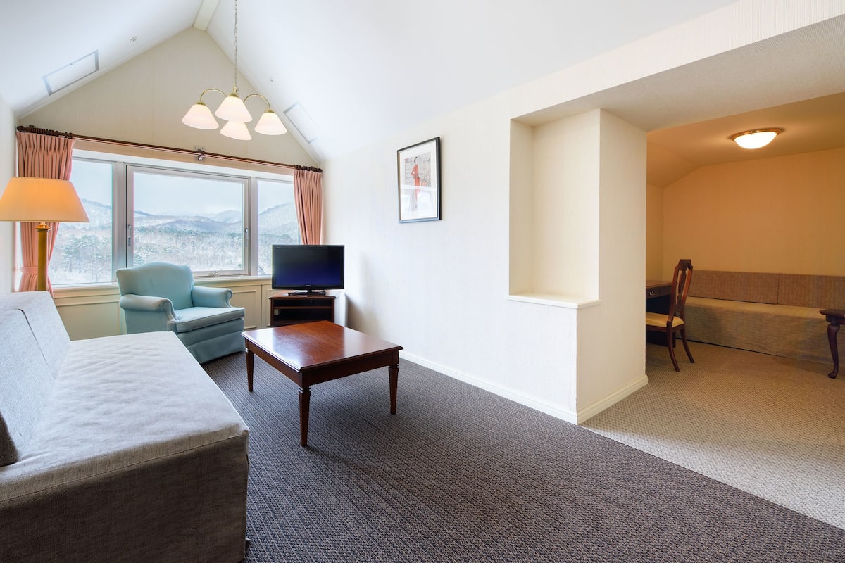 Quarto de hotel com sofá, uma mesinha, televisão, janela com vista para montanhas e abajur.