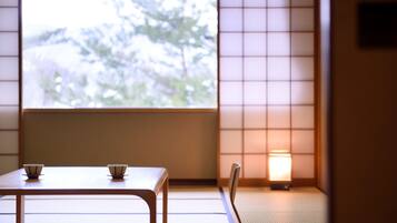 Um espaço com esteiras de tatame, uma mesa baixa e almofadas, uma janela com vista para árvores e um tradicional candeeiro japonês.