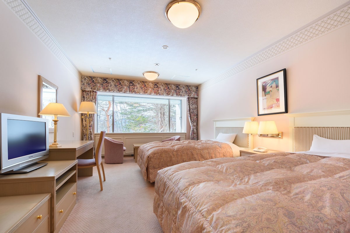 Quarto de hotel com duas camas, uma escrivaninha com televisão e janela com vista para árvores.