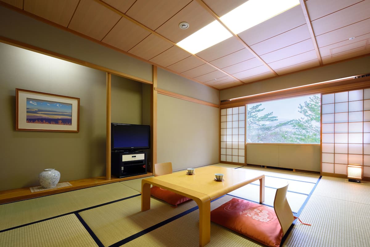 Um quarto japonês tradicional com tatames, uma mesa de madeira baixa, uma televisão e uma janela com vista para árvores.