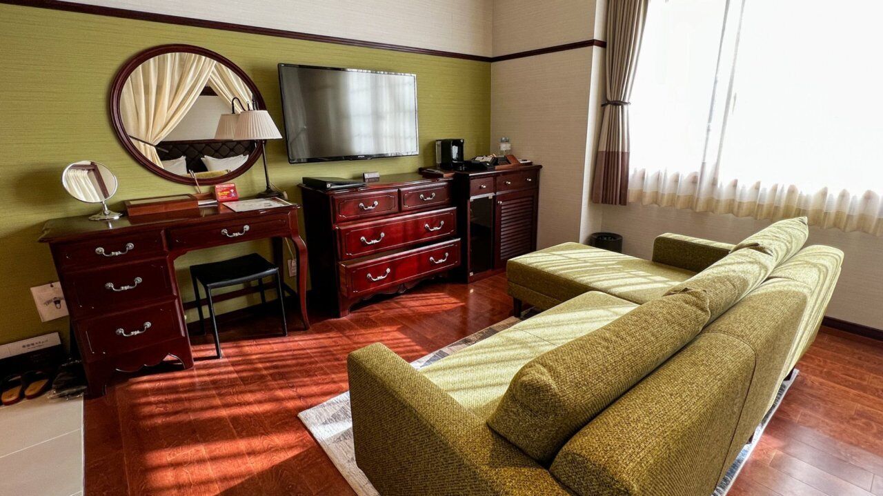 Quarto de hotel com sofá verde, escrivaninha de madeira com gavetas, televisão de tela plana e espelho.