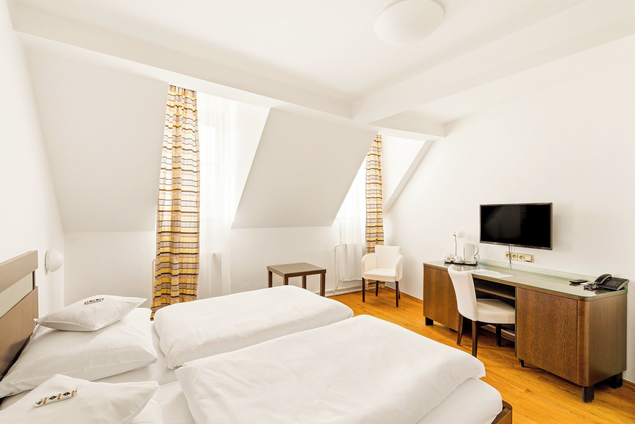Quarto de hotel com duas camas, uma escrivaninha, uma cadeira, televisão e uma janela com cortinas.