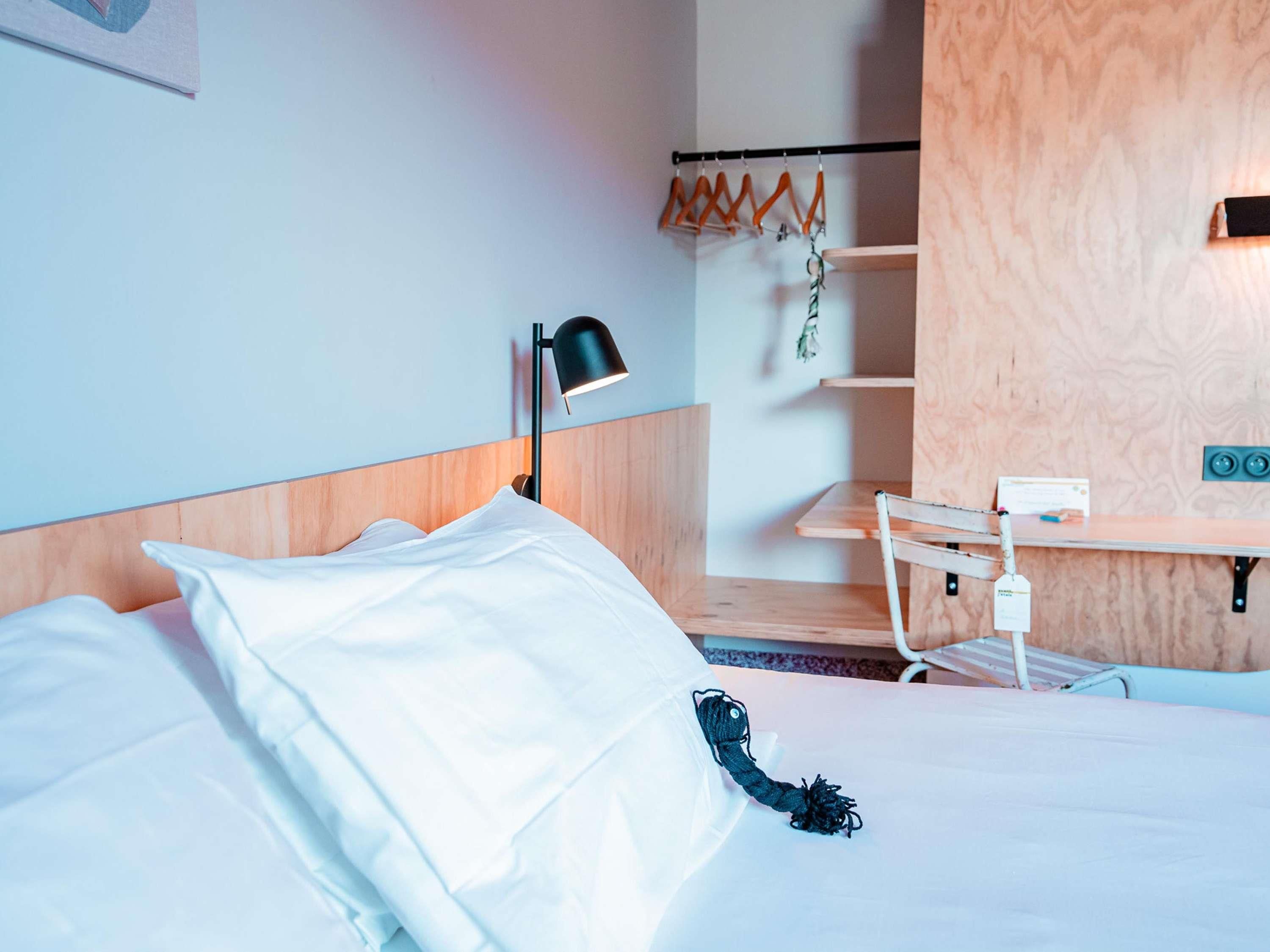 Quarto de hotel com cama, escrivaninha, luminária e prateleira na parede com cabides.