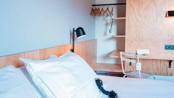 Quarto de hotel com cama, escrivaninha, luminária e prateleira na parede com cabides.