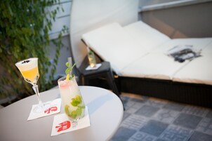 Uma mesa com duas bebidas e um guardanapo com um logotipo, uma cama com um travesseiro e uma revista, e uma planta ao fundo.