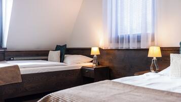 Quarto de hotel com duas camas, cabeceira de madeira, mesa de cabeceira com abajur e janela com persianas.