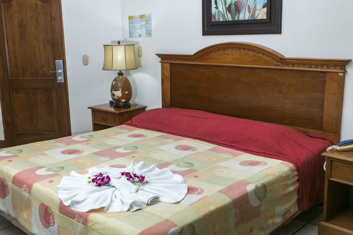 Quarto de hotel com cabeceira de madeira, cama com colcha floral, manta vermelha, mesa de cabeceira com abajur e guarda-roupa de madeira.