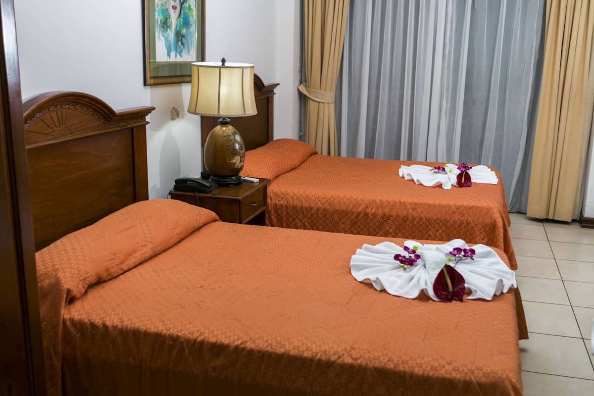 Quarto de hotel com duas camas, cada uma com colcha laranja e toalha branca com flores roxas. Há uma cabeceira de madeira, uma mesa de cabeceira com abajur e um quadro na parede.
