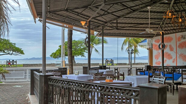 Um restaurante ao ar livre com mesas e cadeiras, vista para o mar e parede decorativa com motivos de folhas de palmeira.