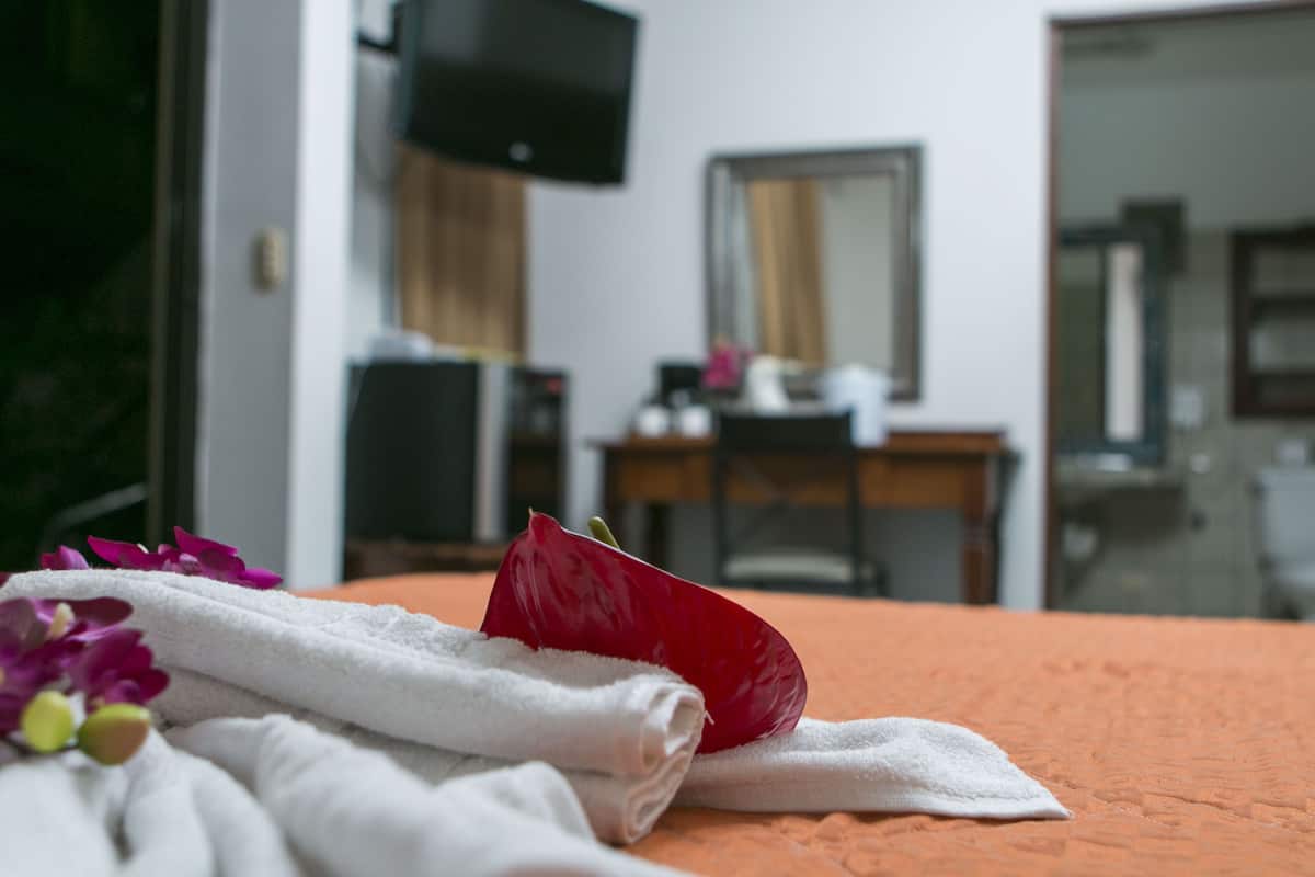 Cama bem arrumada com um objeto decorativo vermelho e toalhas brancas, televisão montada na parede e um espelho sobre uma mesa de madeira.