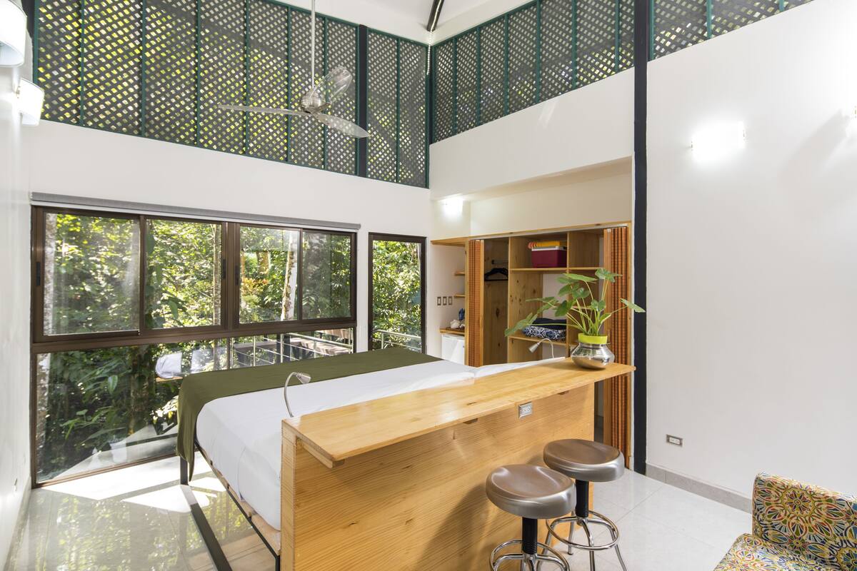 Quarto moderno com uma janela grande, uma escrivaninha de madeira, dois bancos altos e um guarda-roupa.