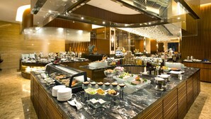 Um saguão de hotel moderno com uma recepção de mármore, uma área de buffet com paredes de vidro e um amplo espaço de estar com detalhes em madeira.