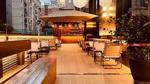 Uma área externa com bancos e mesas de madeira, um bar com placas iluminadas e uma grande planta em vaso.