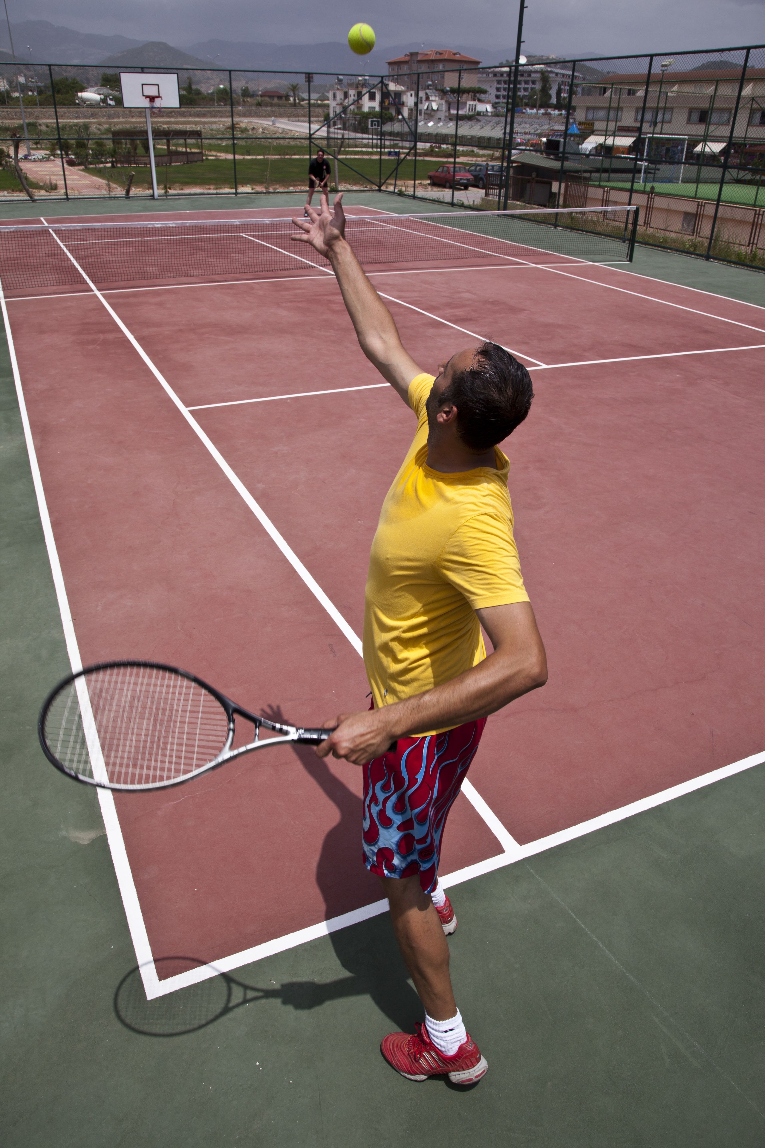 Uma quadra de tênis com uma rede, um jogador vestindo uma camisa amarela e shorts estampados, e uma bola de tênis no ar.