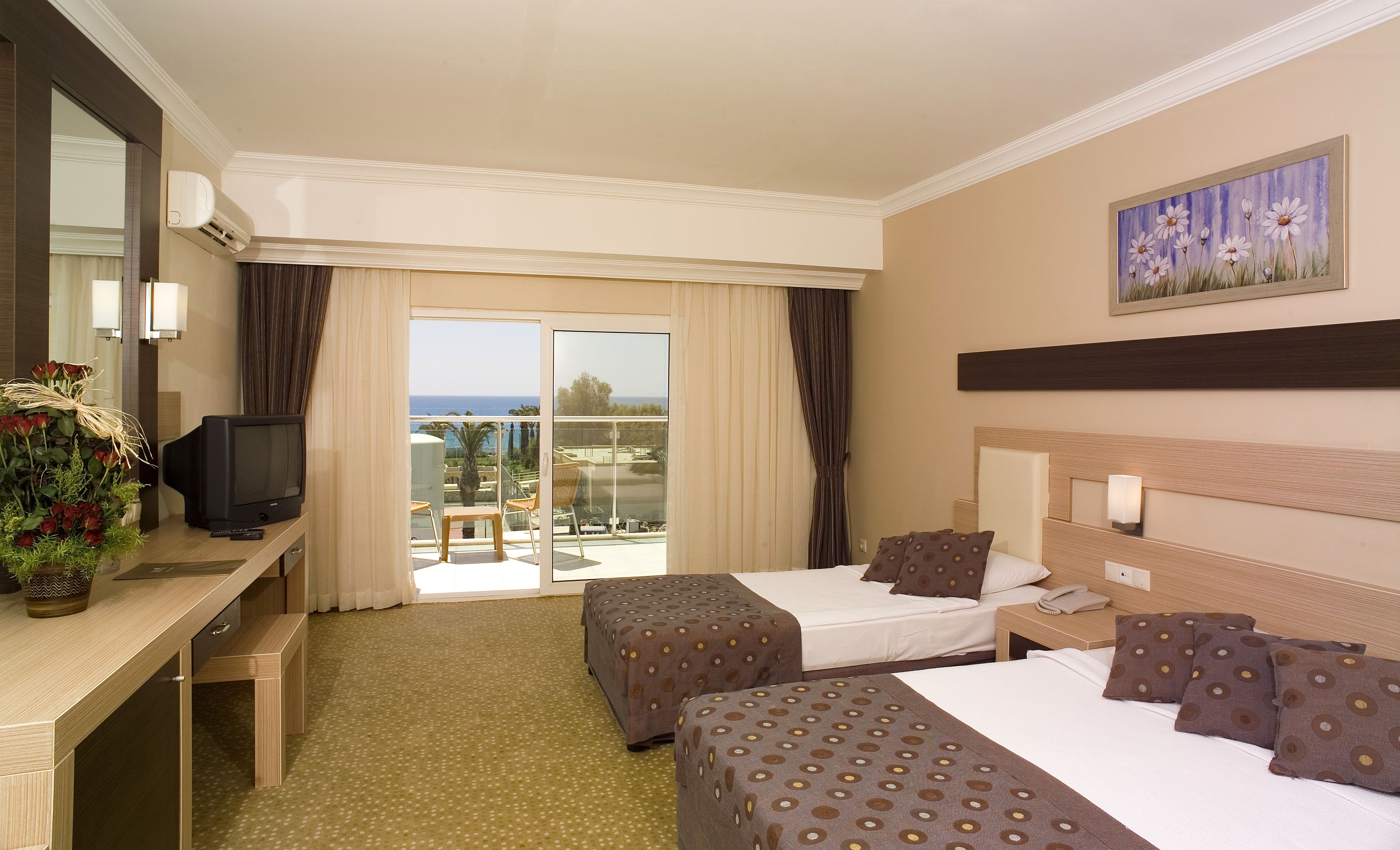 Quarto de hotel com duas camas, uma escrivaninha com televisão, uma janela grande com vista para o mar e um quadro na parede.
