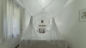 Quarto com cama sob um dossel branco, uma mesinha com toalha de mesa florida e um quadro na parede.