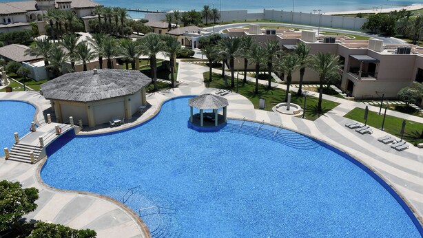 Uma piscina grande com um gazebo, palmeiras e vista para o horizonte da cidade e praia.