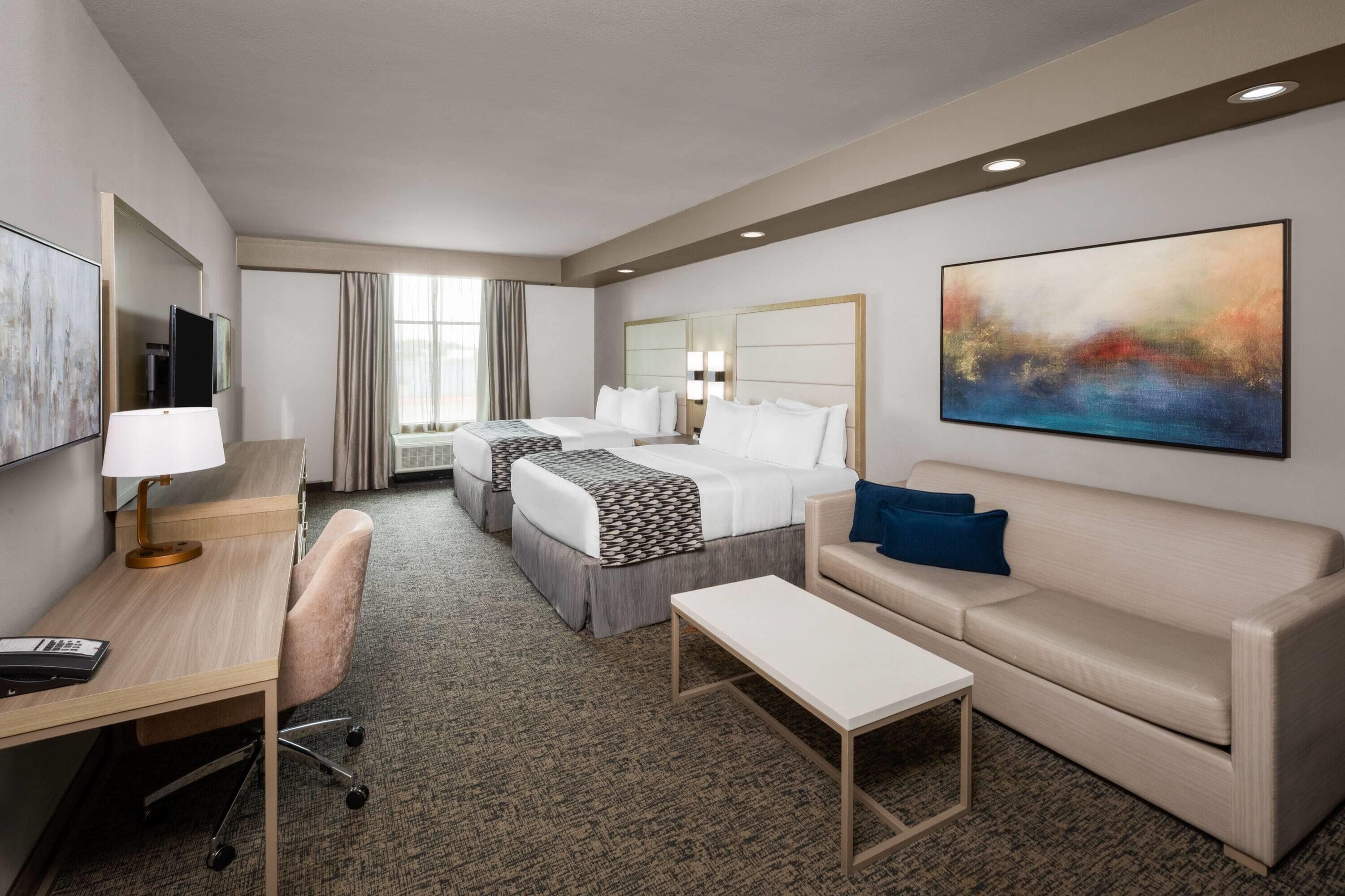 Quarto de hotel com duas camas, uma escrivaninha com luminária, um sofá, uma mesa de centro e uma grande pintura abstrata na parede.