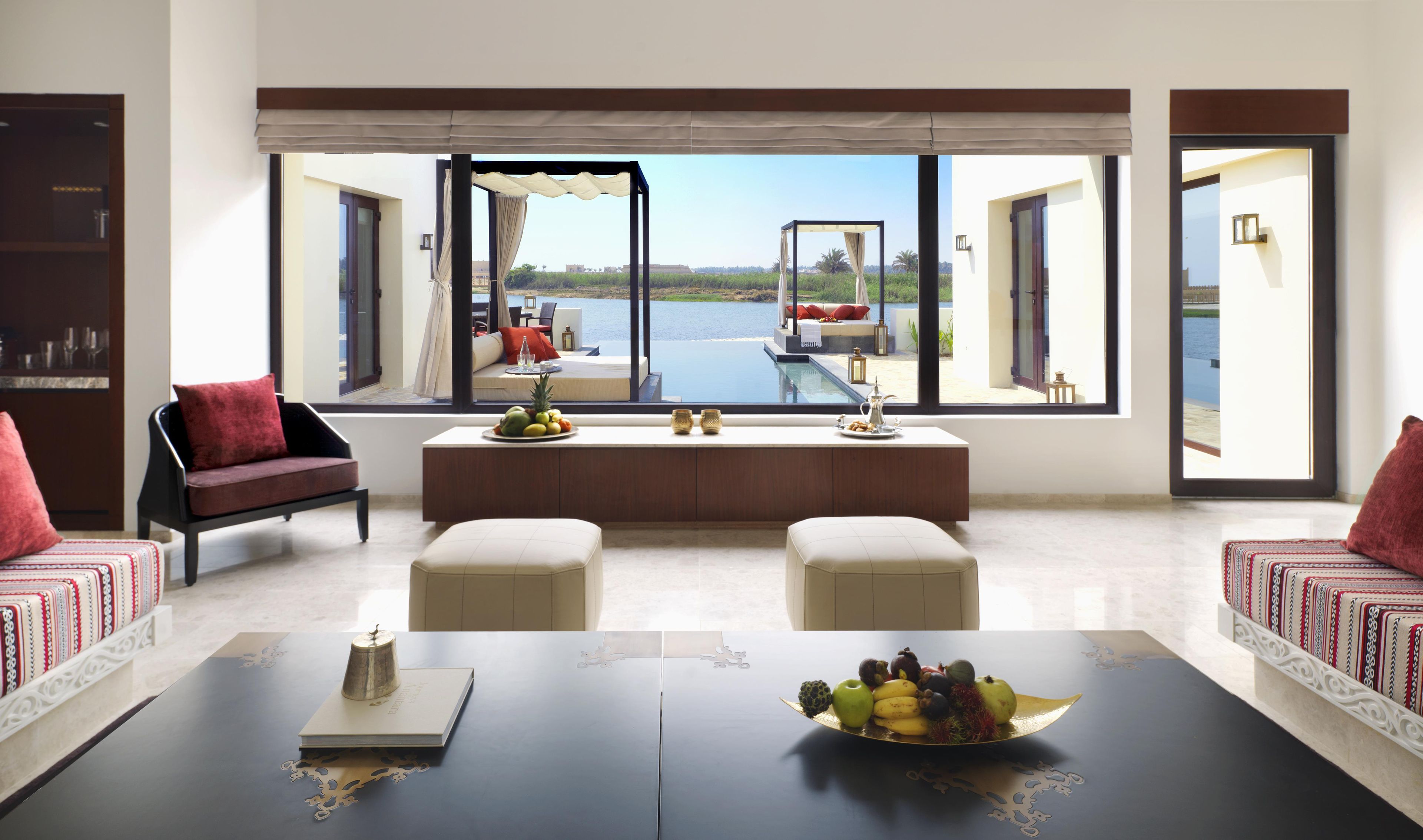 Sala de estar moderna com uma grande janela que dá para uma piscina e uma vista panorâmica. Há um sofá, uma mesa de centro com frutas e uma estante de livros.