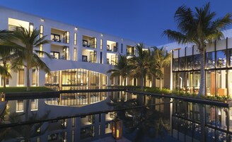 Um hotel moderno com várias varandas, uma piscina espelhada e palmeiras.