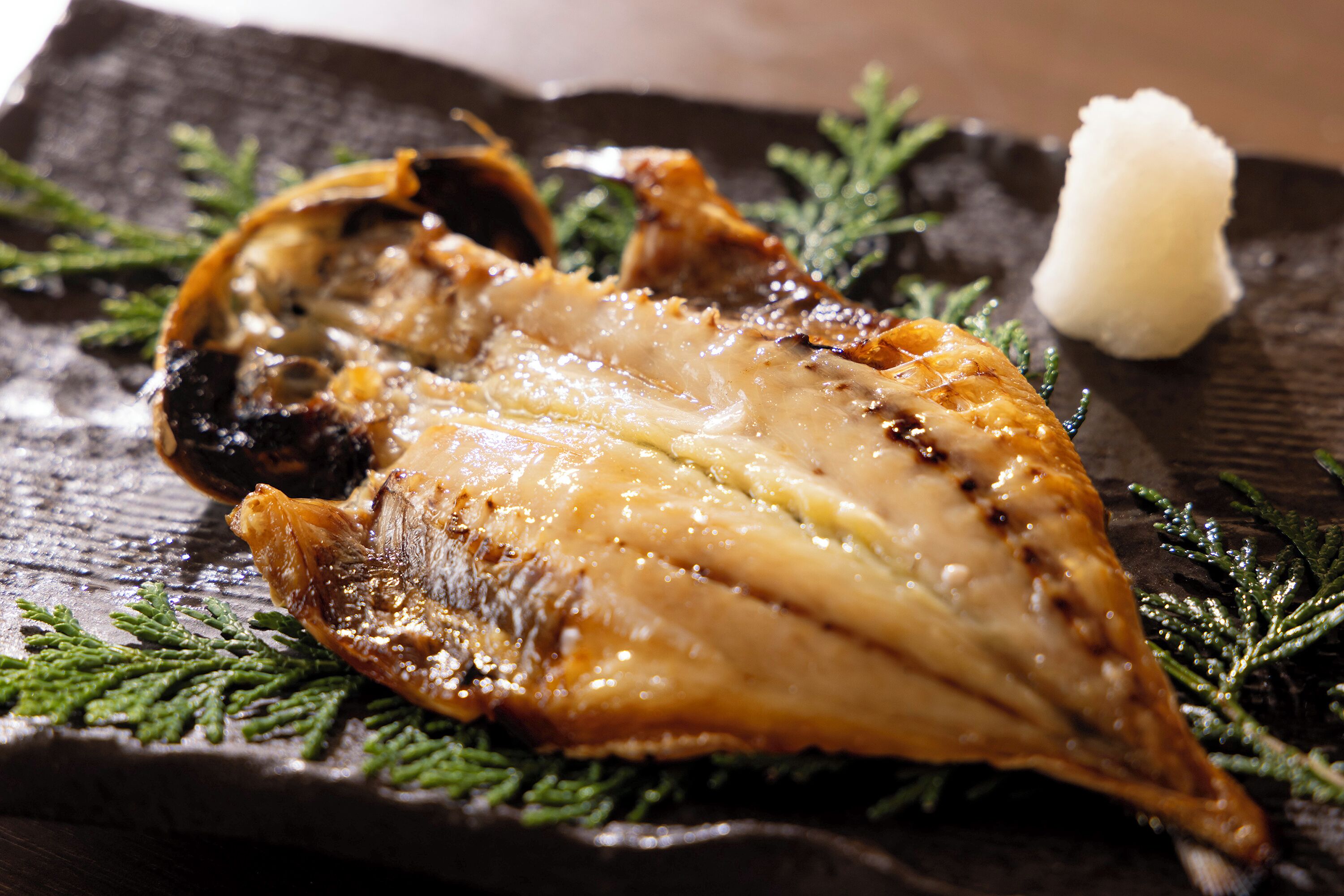 Close-up de um peixe cozido com pele crocante, guarnecido com ervas verdes e uma pequena porção de arroz em um prato escuro.