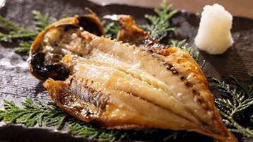 Close-up de um peixe cozido com pele crocante, guarnecido com ervas verdes e uma pequena porção de arroz em um prato escuro.