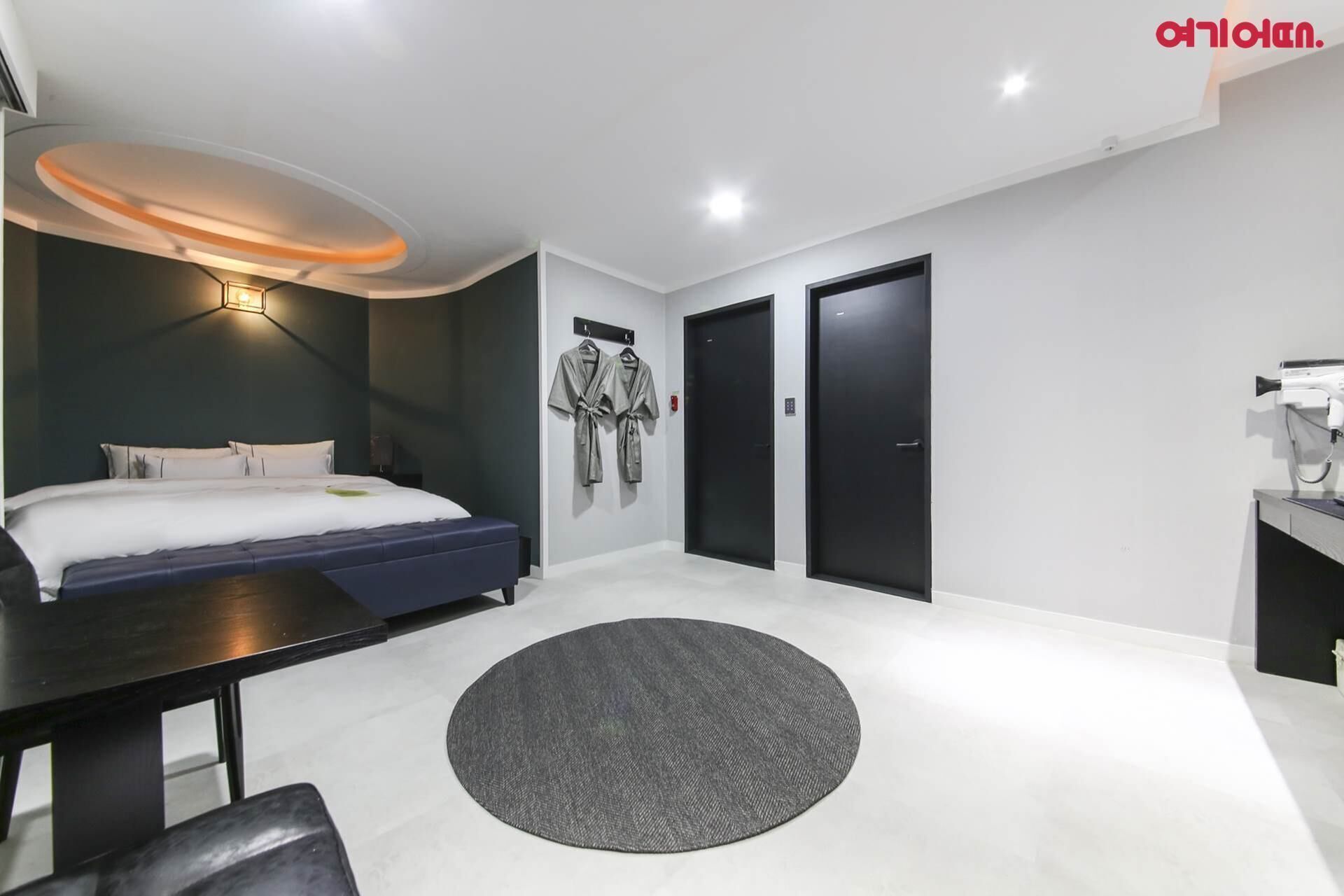 Quarto de hotel moderno com uma cama, uma escrivaninha, um tapete redondo e duas portas pretas.