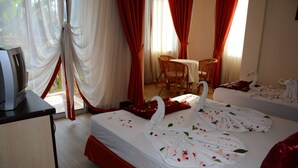 Quarto de hotel com duas camas, televisão, área de refeições com mesa e cadeiras, e cortinas vermelhas.
