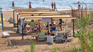 Uma praia com espreguiçadeiras e guarda-sóis sob área sombreada, um letreiro com a inscrição "YAVUZHAN OTEL BEACH & STRAND" e pessoas nadando na água.