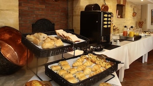 Buffet com diversos pães, incluindo pão, croissants e doces, exposto numa mesa com uma parede de tijolos e panelas de cobre ao fundo.
