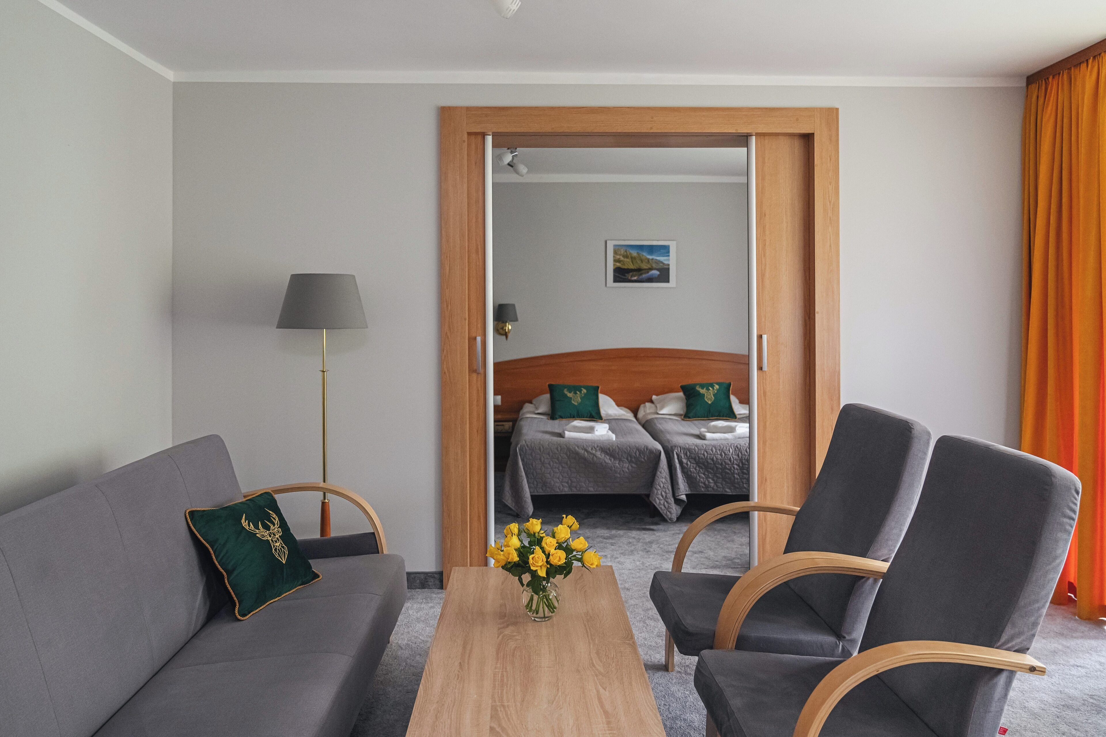 Quarto de hotel com sofá, poltrona e uma pequena mesa com um vaso de flores. Há duas camas com almofadas verdes e um quadro na parede.