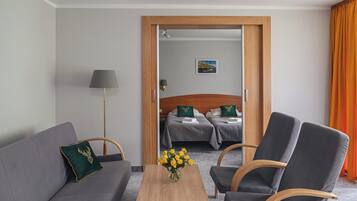 Quarto de hotel com sofá, poltrona e uma pequena mesa com um vaso de flores. Há duas camas com almofadas verdes e um quadro na parede.