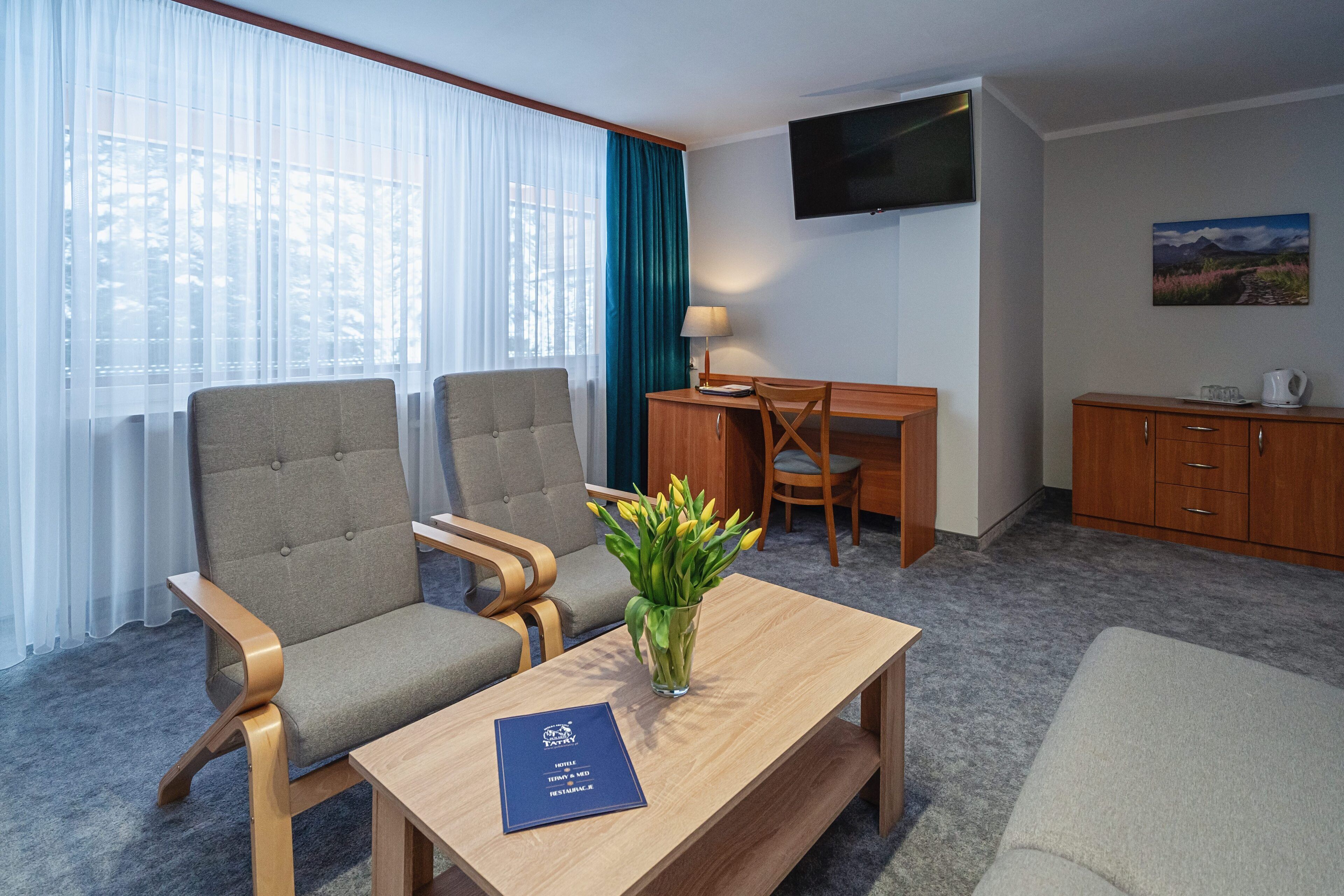 Quarto de hotel com duas poltronas, uma mesa de centro de madeira com um livro, uma escrivaninha com abajur, TV na parede e um quadro.