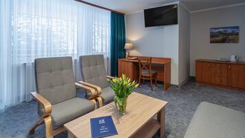 Quarto de hotel com duas poltronas, uma mesa de centro de madeira com um livro, uma escrivaninha com abajur, TV na parede e um quadro.