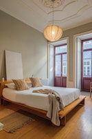 Quarto com cama de madeira, luminária pendente e vista para o exterior por grandes janelas.