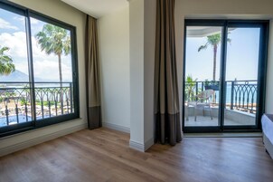 Quarto com grandes janelas, varanda com vista para a praia e palmeiras, e piso de madeira.