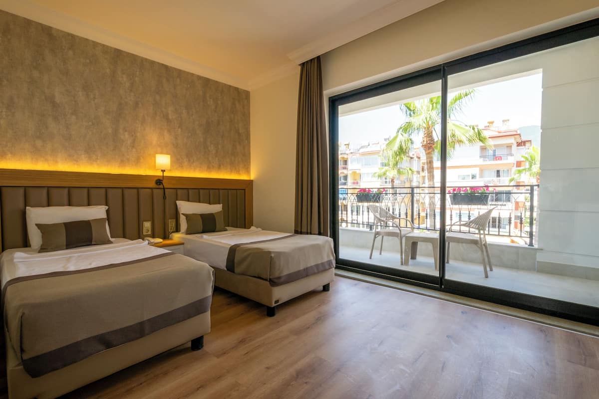 Quarto de hotel com duas camas, uma janela grande com vista para palmeiras e edifícios, e uma varanda com cadeiras.