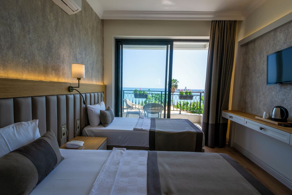 Quarto de hotel com duas camas, televisão, varanda com vista para o mar e janela com cortinas.