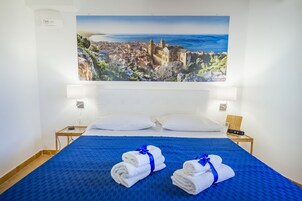 Quarto de hotel com uma cama grande, duas almofadas brancas, um edredom azul e duas toalhas enroladas com fitas azuis.