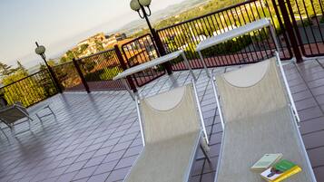 Terraço com duas cadeiras de descanso e uma mesa, com vista para uma paisagem cênica com edifícios e vegetação.