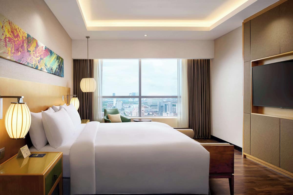 Quarto de hotel com uma cama grande, uma escrivaninha com abajur, televisão e vista para a cidade pela janela.