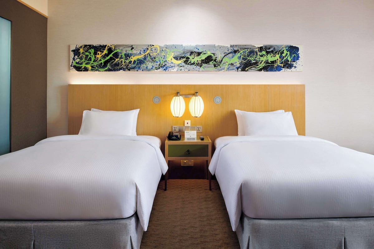 Quarto de hotel com duas camas, cabeceira de madeira, mesa de cabeceira com abajur e uma pintura abstrata moderna acima da cabeceira.