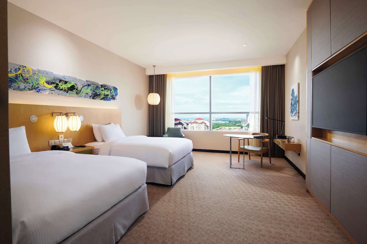 Quarto de hotel com duas camas, uma escrivaninha, uma cadeira, uma televisão e uma janela grande com vista.