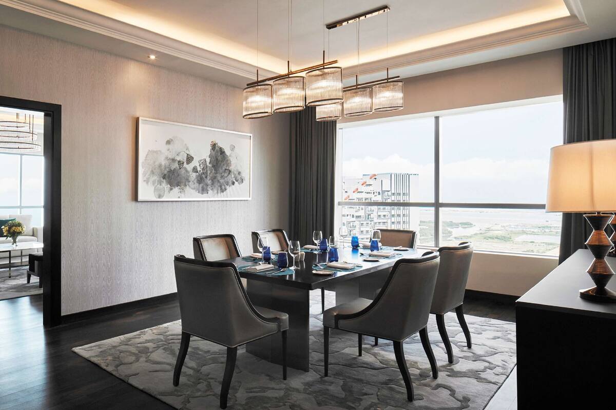 Quarto de hotel moderno com área de refeições, conjunto de sofás e uma grande janela com vista para a cidade.
