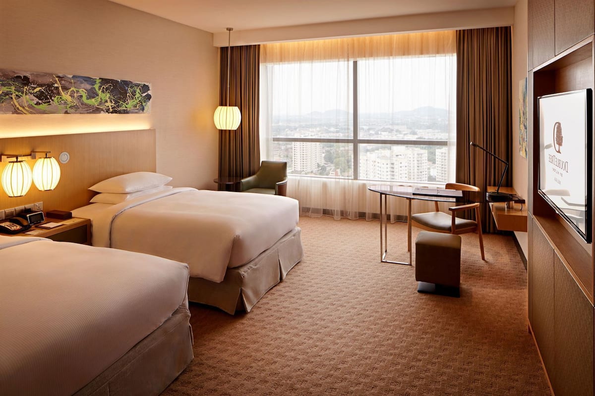 Quarto de hotel com duas camas, uma escrivaninha, uma cadeira, uma televisão e uma janela grande com vista para a cidade.