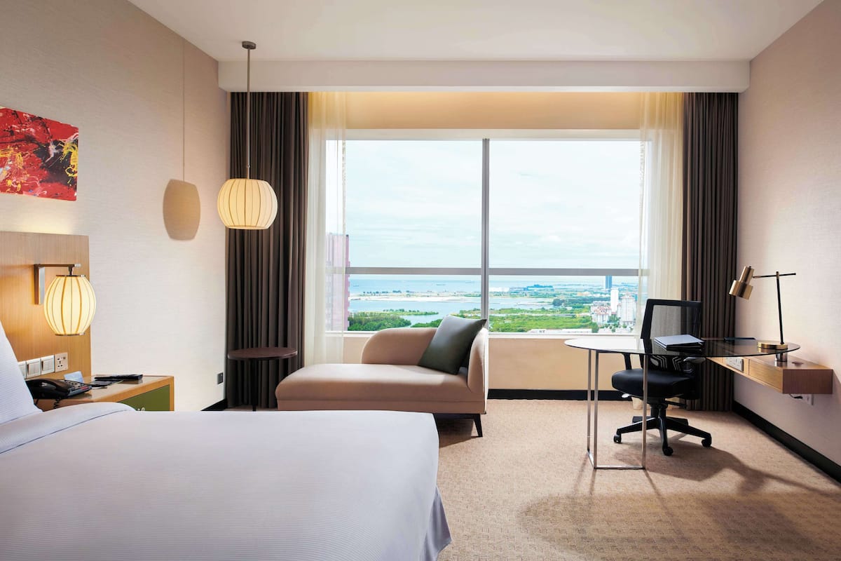 Quarto de hotel com cama, escrivaninha, cadeira, luminária, televisão e uma grande janela com vista.