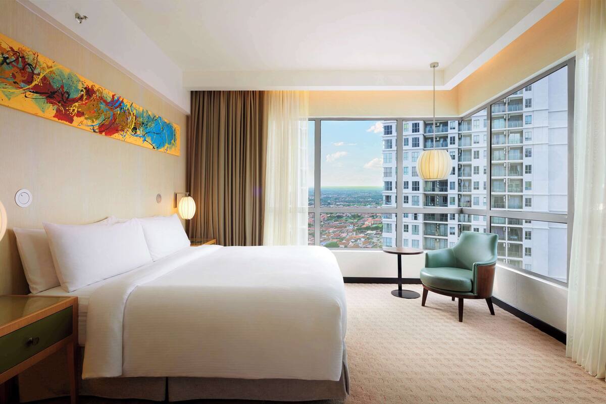Quarto de hotel com uma cama grande, vista para a paisagem urbana, poltrona verde e uma pintura abstrata colorida na parede.
